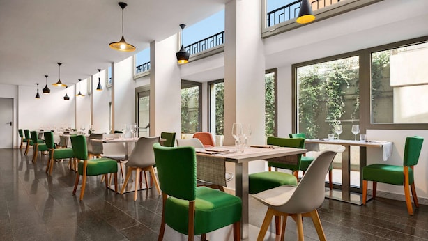 Uma sala de jantar ampla, com várias mesas e cadeiras, grandes janelas e iluminação pendente.