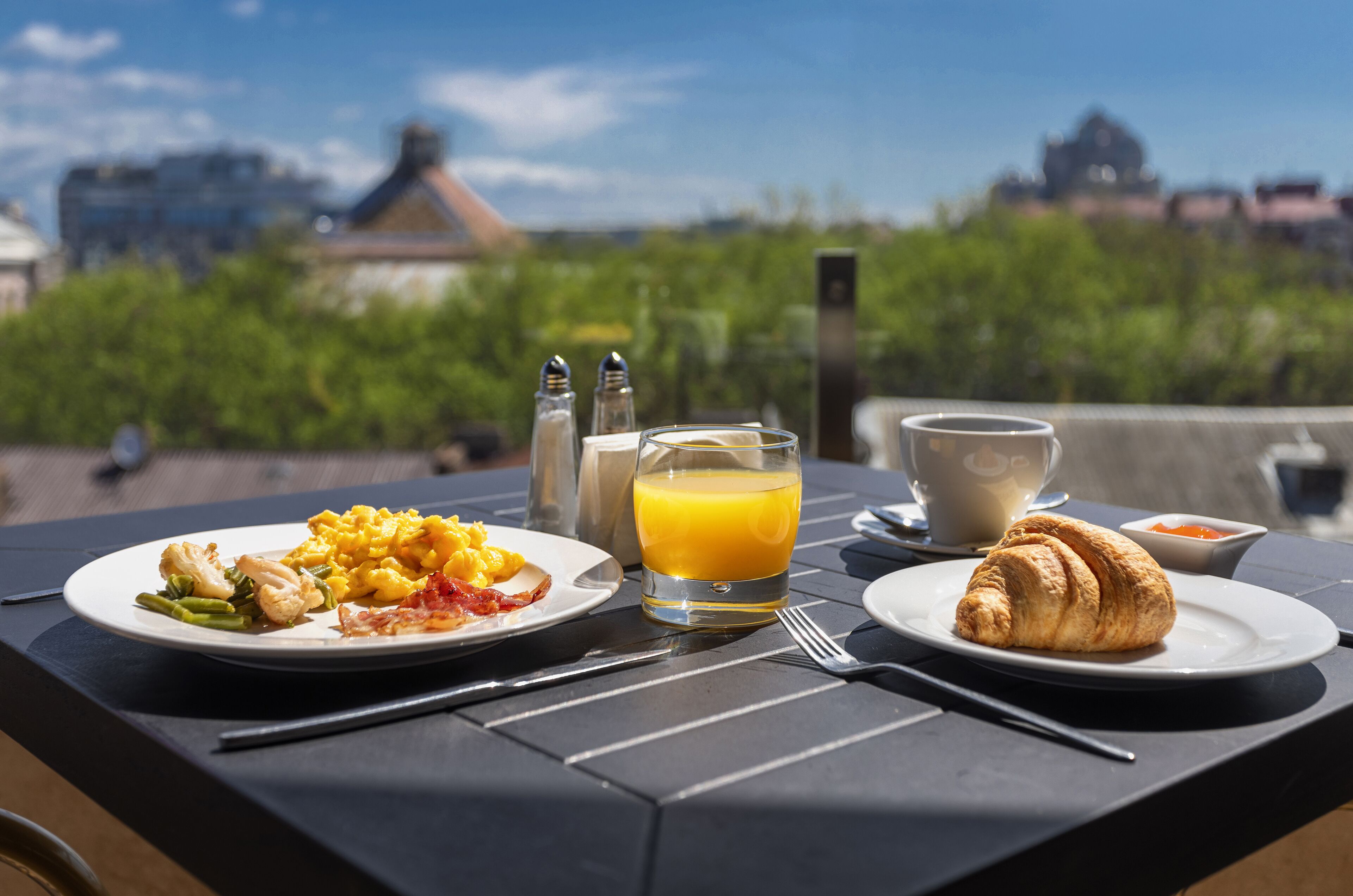 Uma mesa com itens de café da manhã, incluindo ovos mexidos, torradas e um croissant, além de um copo de suco de laranja e uma xícara de café.