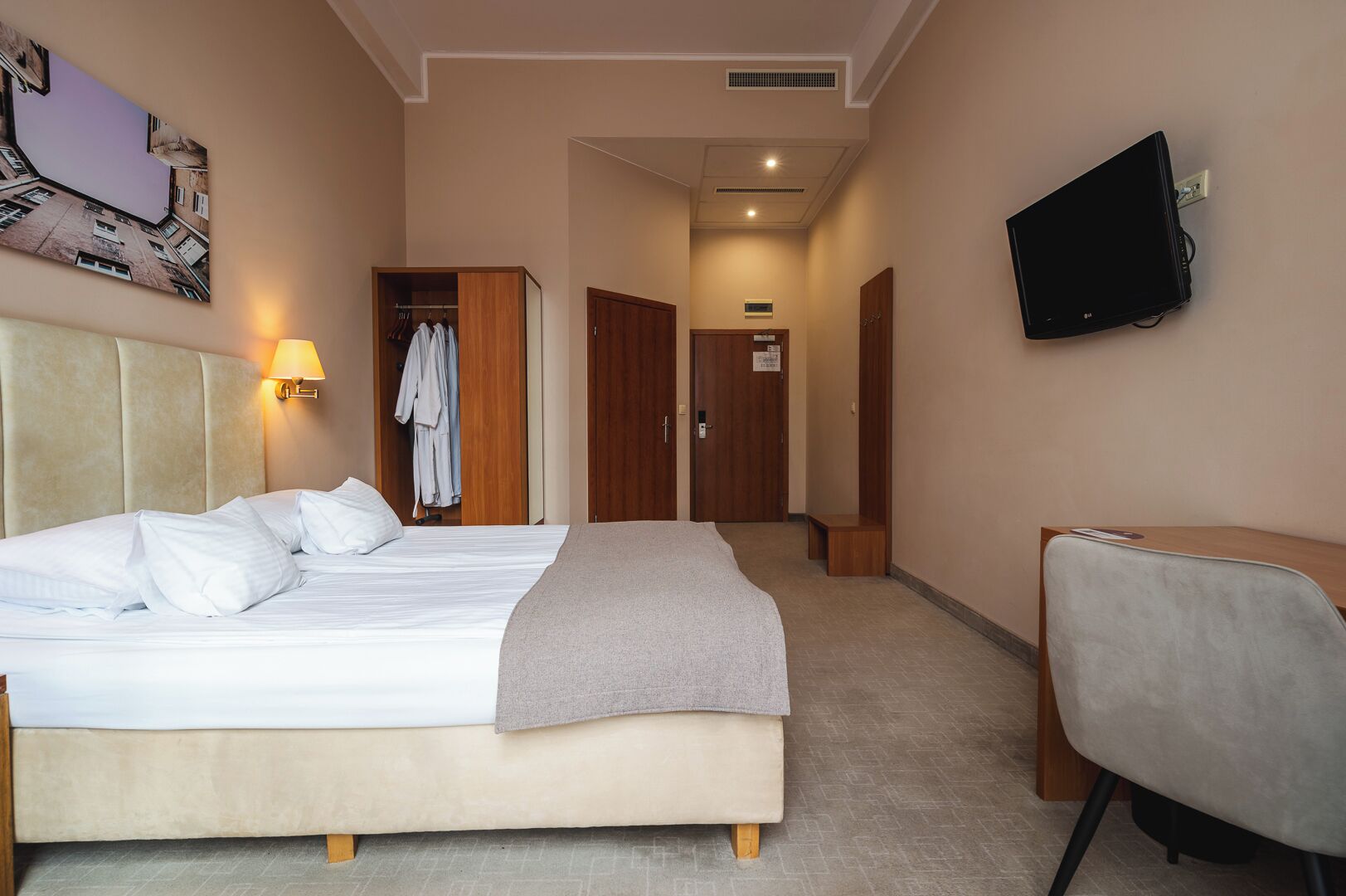 Quarto de hotel com cama, escrivaninha, cadeira, televisão, guarda-roupa e corredor.