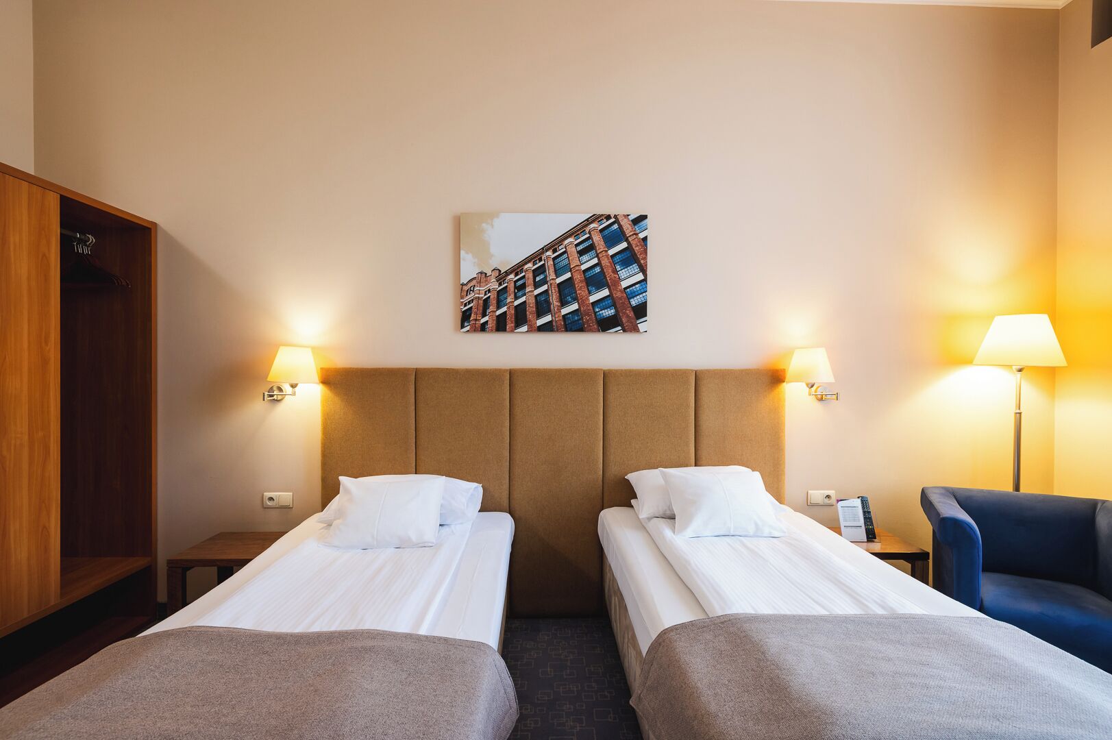 Quarto de hotel com duas camas, um guarda-roupa, uma escrivaninha, uma cadeira e um quadro na parede.