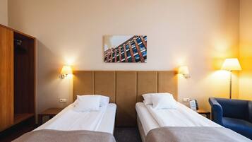 Quarto de hotel com duas camas, um guarda-roupa, uma escrivaninha, uma cadeira e um quadro na parede.