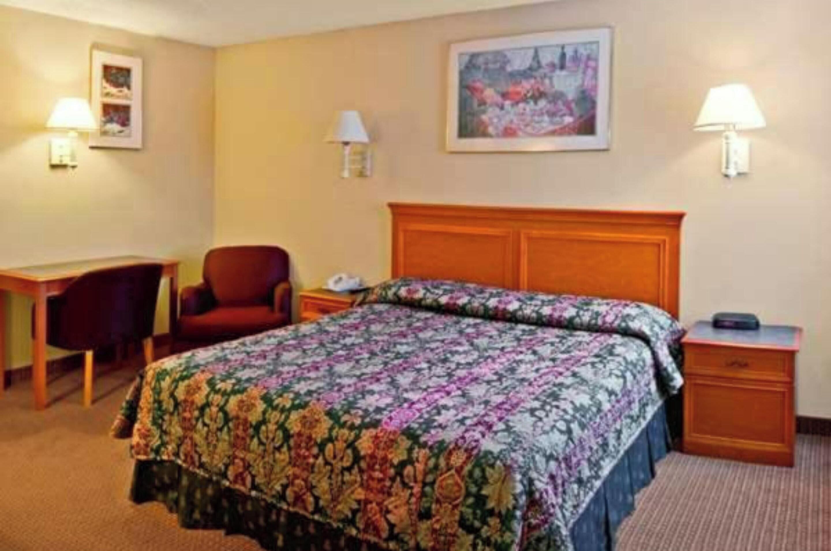 Quarto de hotel com cama, escrivaninha, cadeira, mesa de cabeceira e abajur.