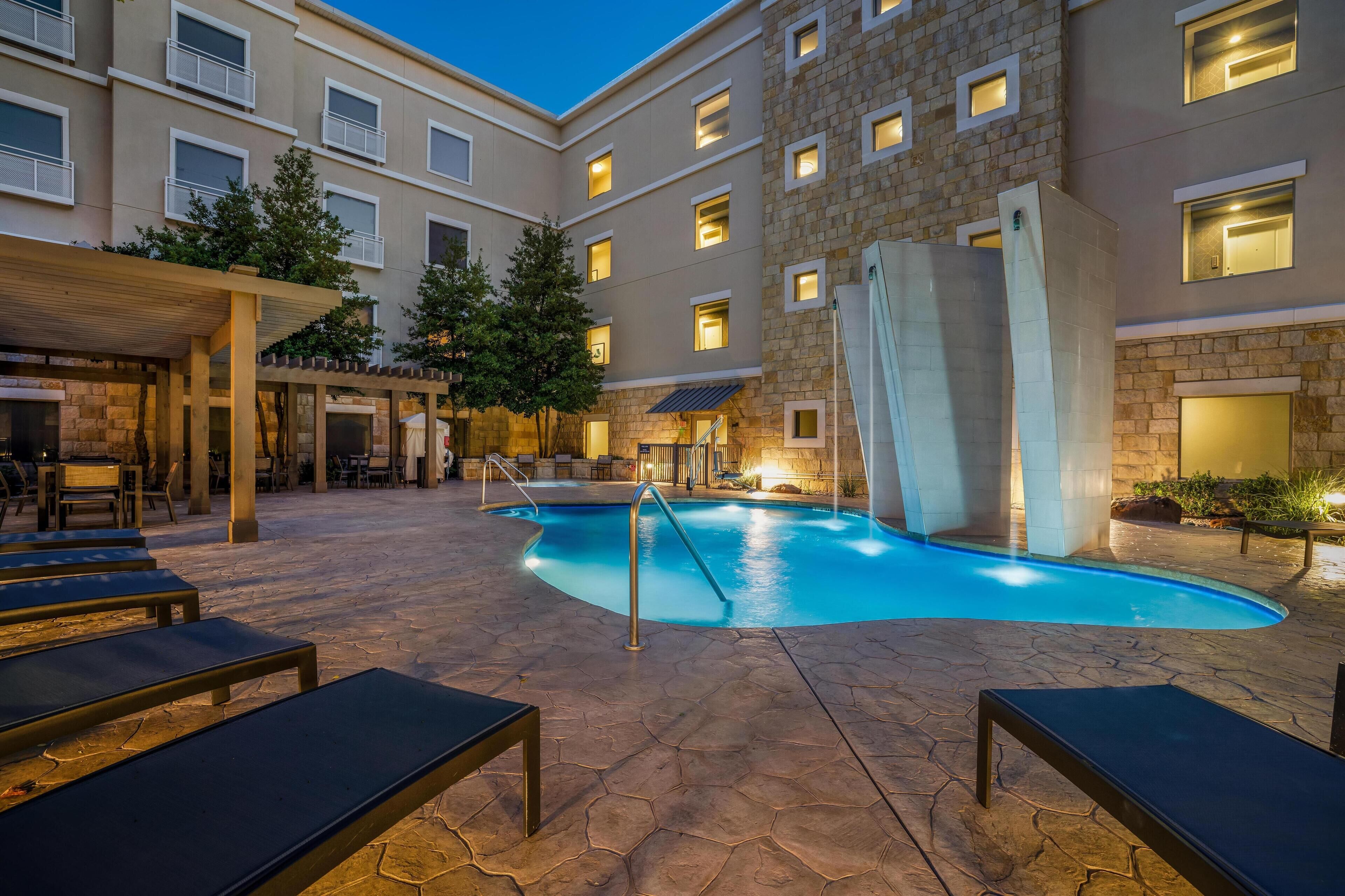 Uma área de piscina com espreguiçadeiras, um espaço de estar coberto e um edifício de pedra com várias janelas.