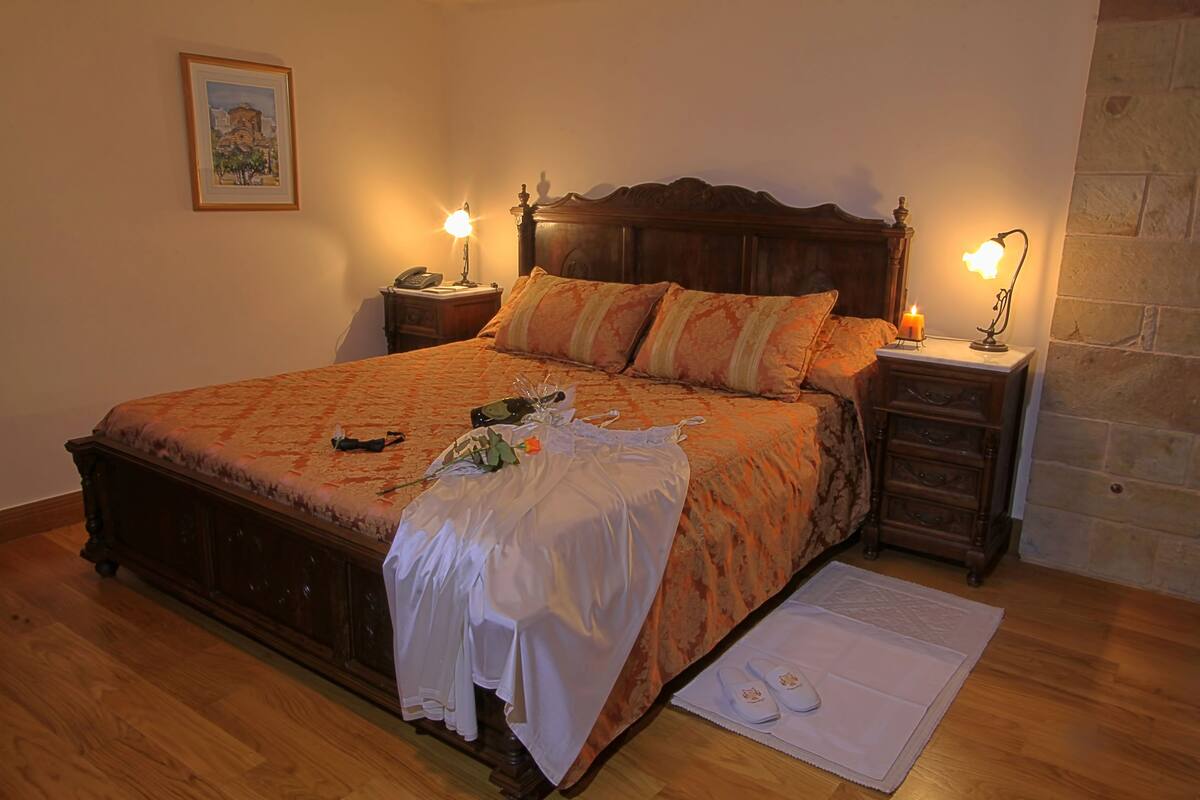 Cama bem arrumada com colcha florida, duas luminárias de cabeceira, mesa de cabeceira com telefone e um quadro na parede.