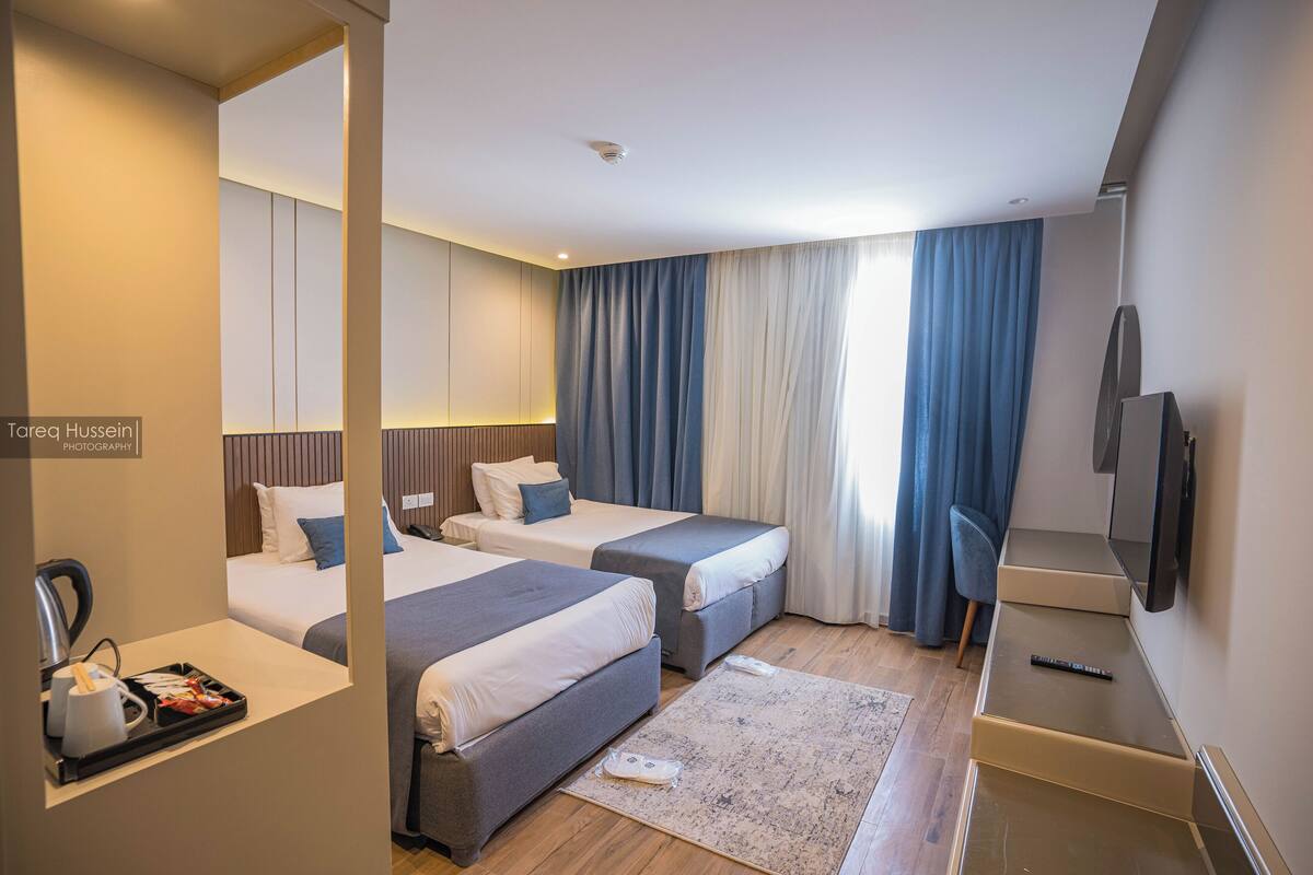 Quarto de hotel com duas camas, uma televisão, uma secretária com uma chaleira e um tabuleiro com comida.