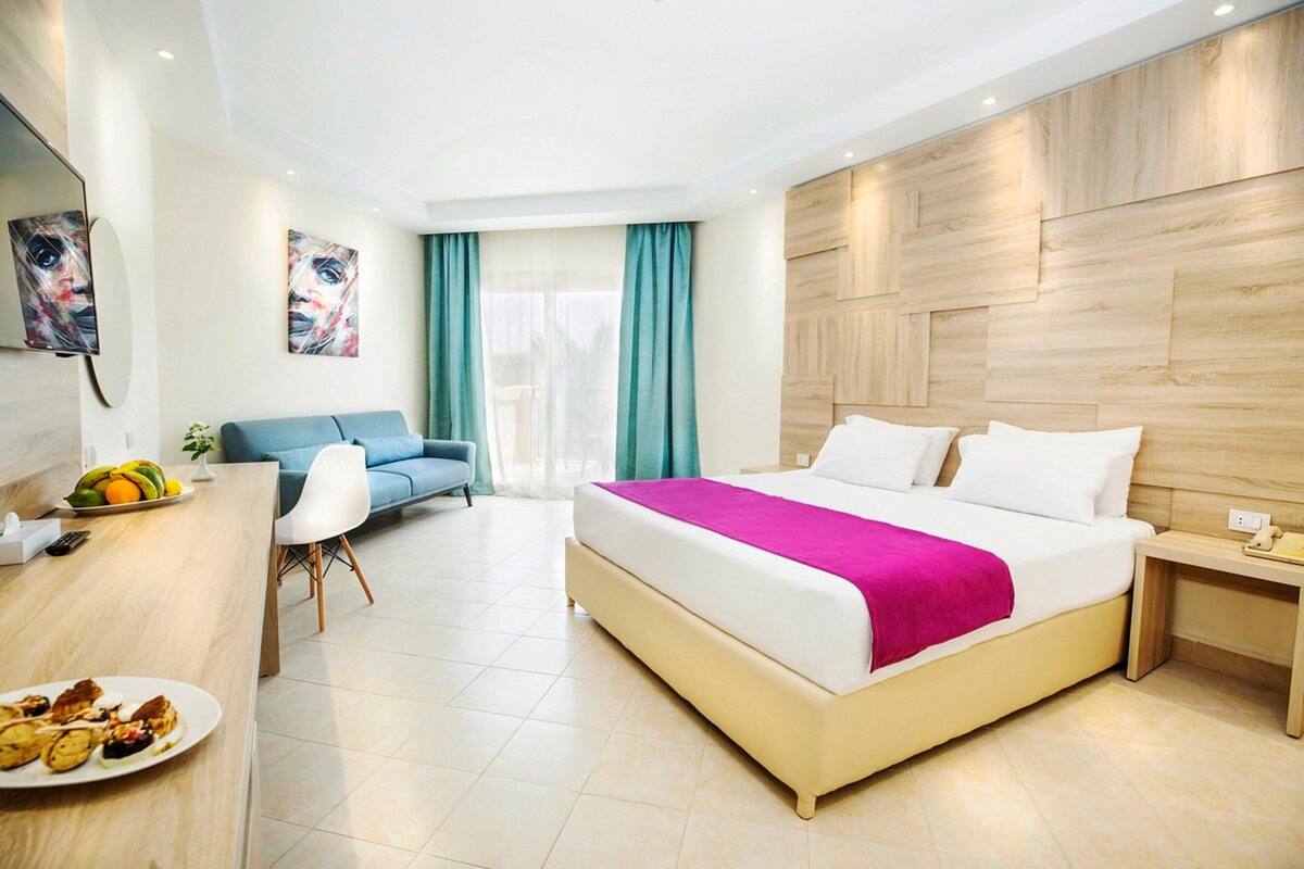 Quarto de hotel moderno com uma cama grande, uma escrivaninha com um prato de comida, uma poltrona azul e uma cabeceira de madeira.