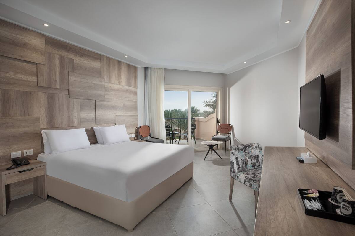 Quarto de hotel moderno com uma cama grande, escrivaninha, cadeira, televisão e varanda com vista para palmeiras.