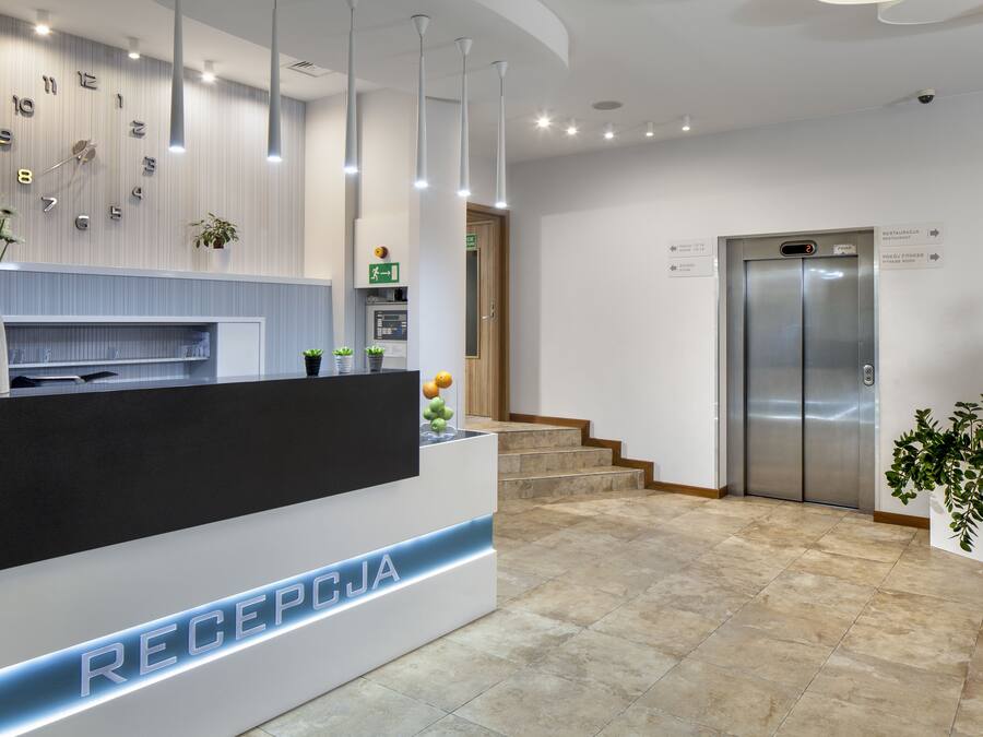 Uma área de recepção com balcão de atendimento identificado como “RECEPTION”, um relógio digital, uma escada que dá acesso a um elevador e um vaso de planta.