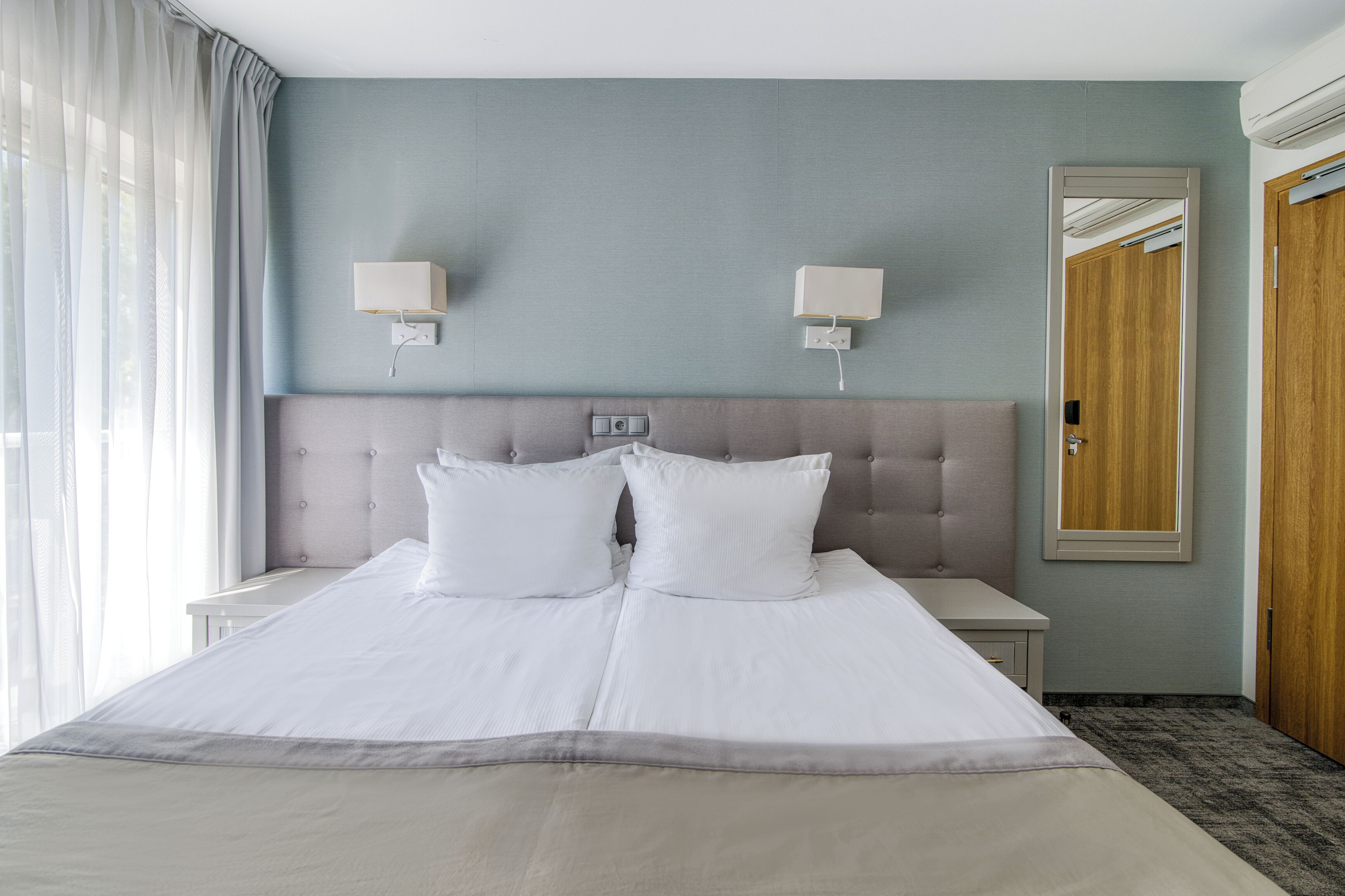 Uma cama de casal bem arrumada com roupa de cama branca, uma cabeceira estofada, duas mesas de cabeceira com luminárias e uma porta de madeira.