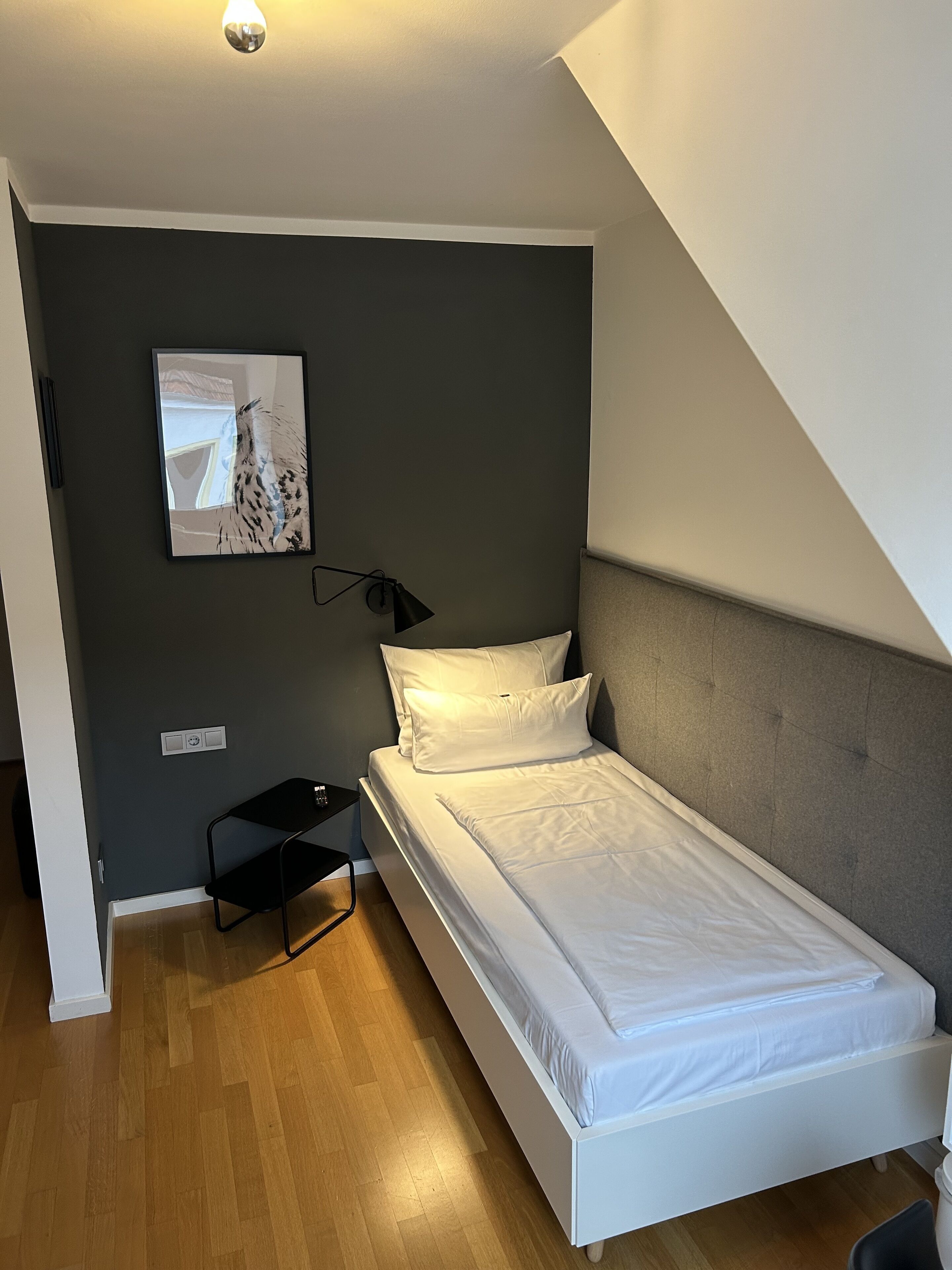 Quarto pequeno com uma cama de solteiro, teto inclinado, mesa de cabeceira, abajur e um quadro na parede.