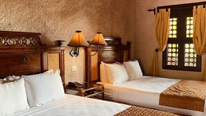 Quarto de hotel com duas camas, cabeceira de madeira, mesa de cabeceira e janela com cortinas.