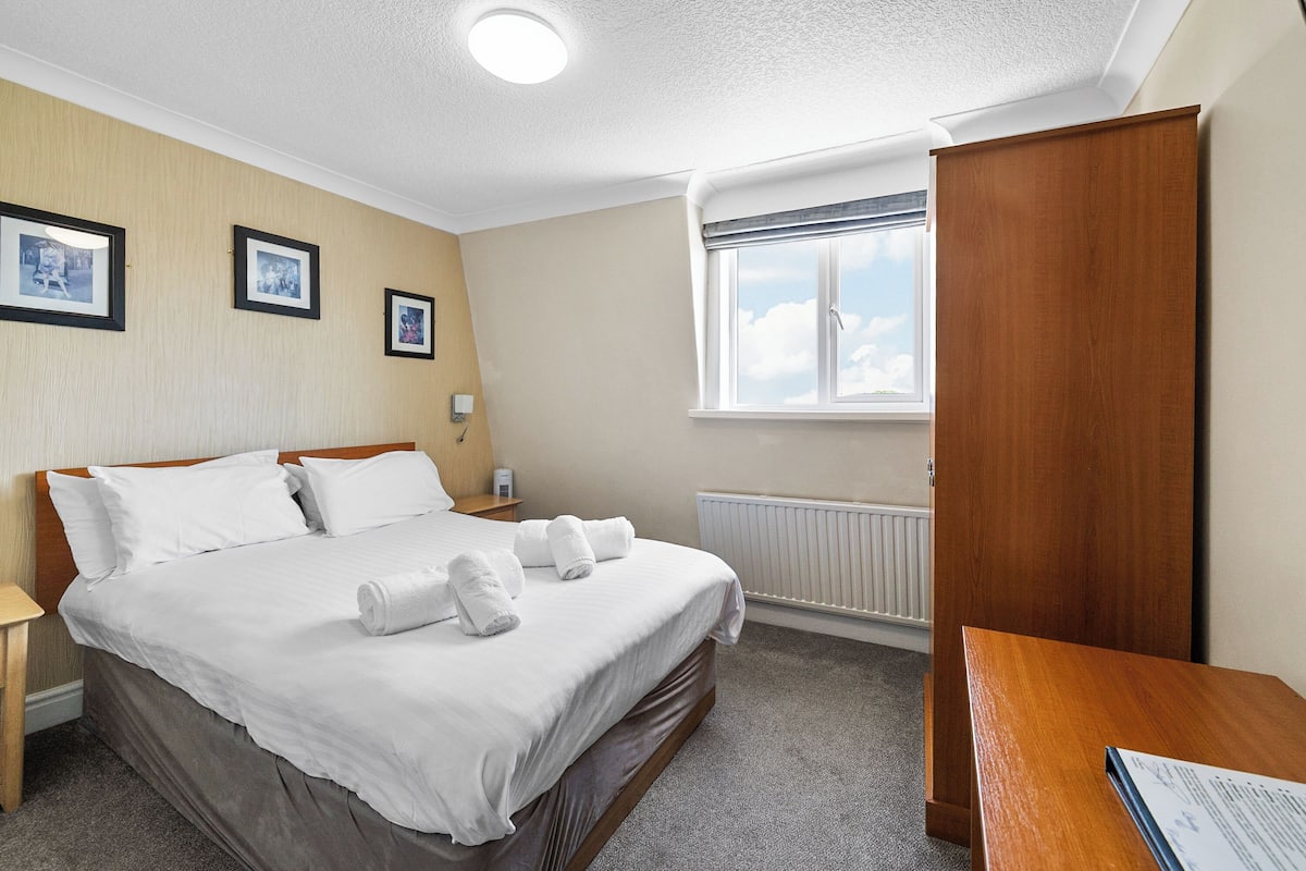 Uma cama bem arrumada com roupa de cama e toalhas brancas, uma cabeceira de madeira, uma mesa de cabeceira, uma janela com vista para o céu e um guarda-roupa de madeira.