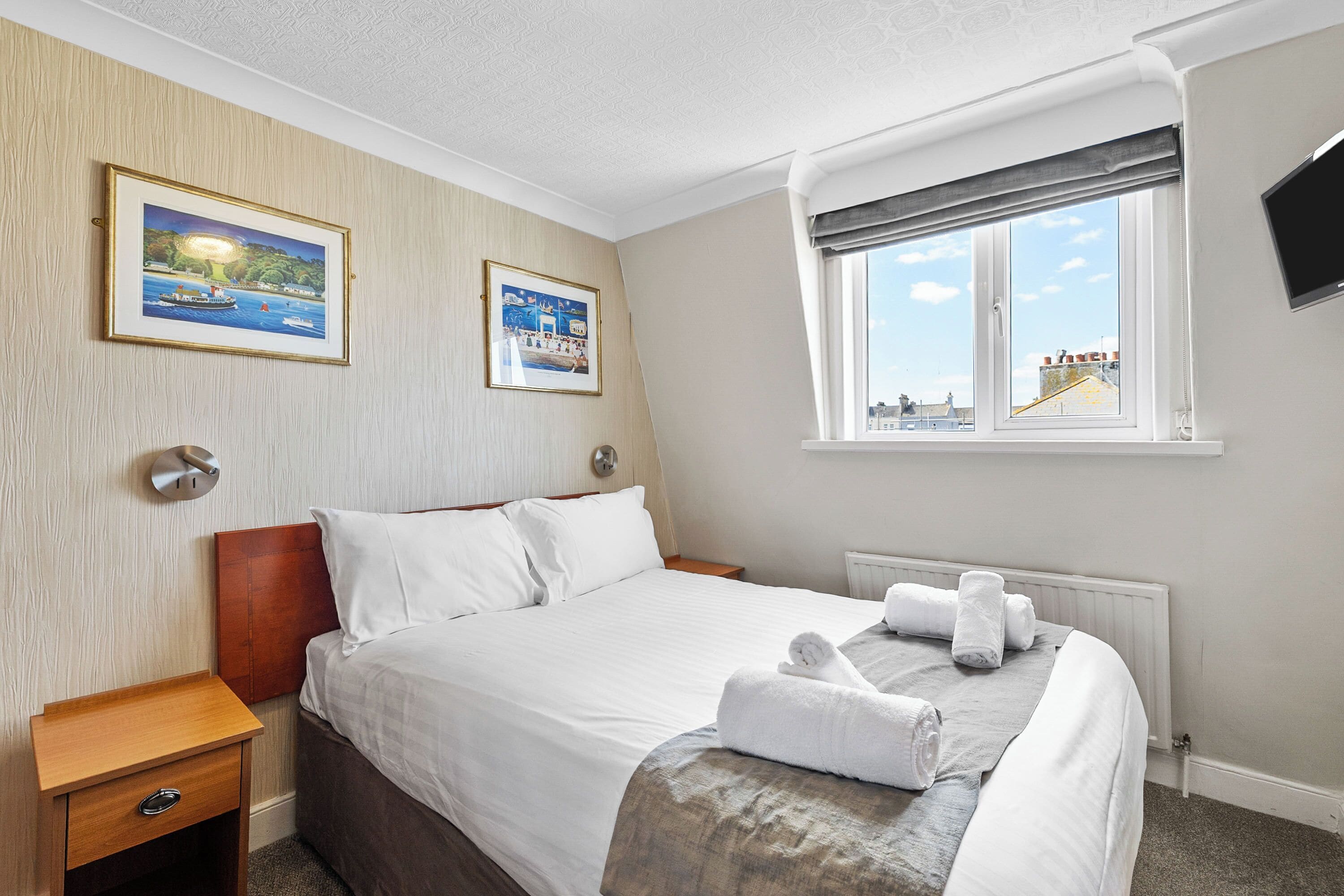 Quarto de hotel com uma cama bem arrumada, uma mesa de cabeceira, uma janela com vista para prédios e duas imagens emolduradas na parede.