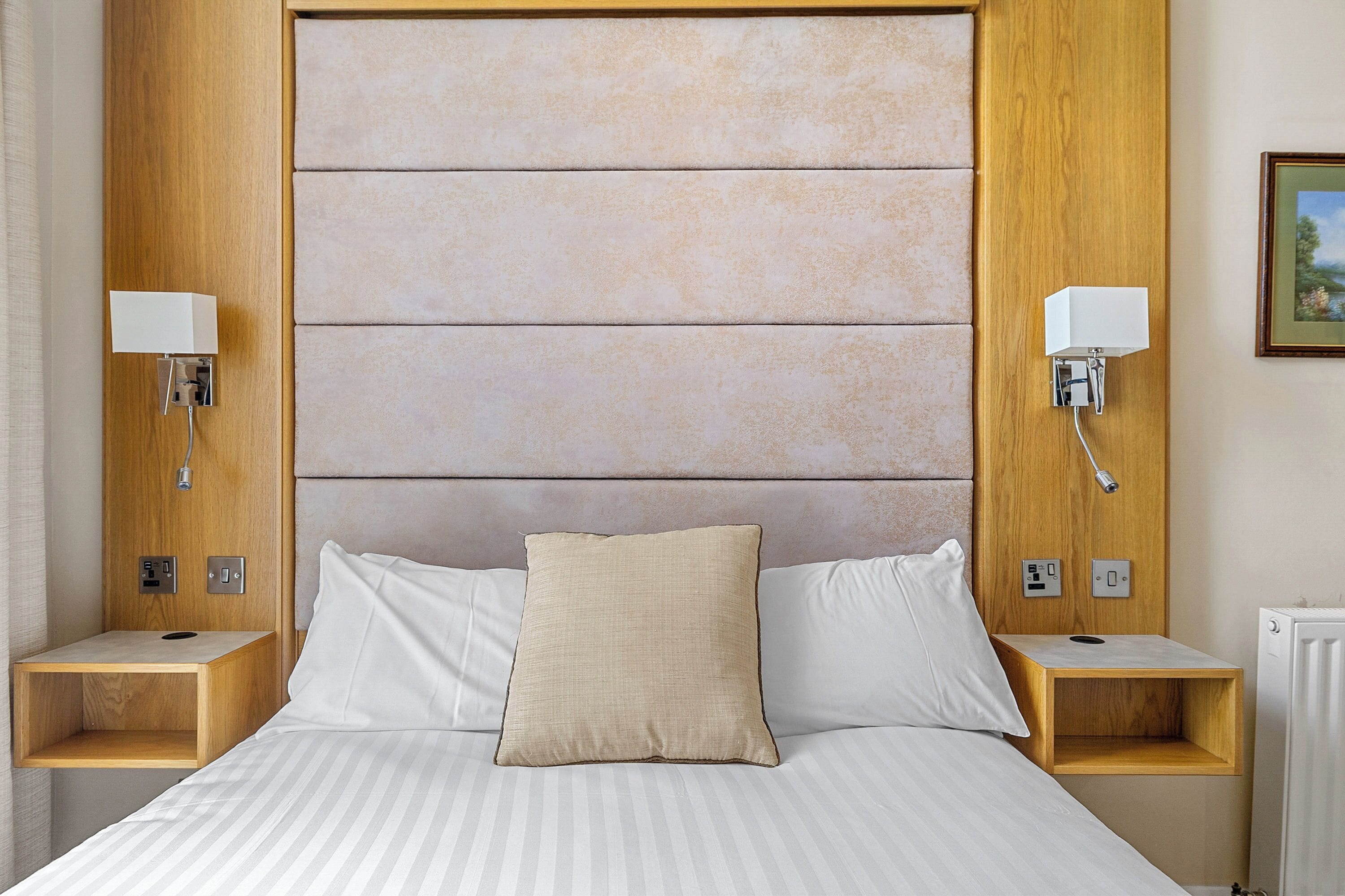 Quarto de hotel com uma cama grande, duas mesas de cabeceira, encosto de cama e luminárias fixadas na parede.