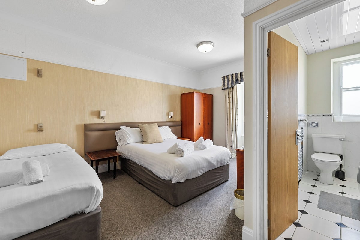Quarto de hotel com duas camas, um guarda-roupa de madeira, uma mesinha de cabeceira e banheiro com vaso sanitário e pia.