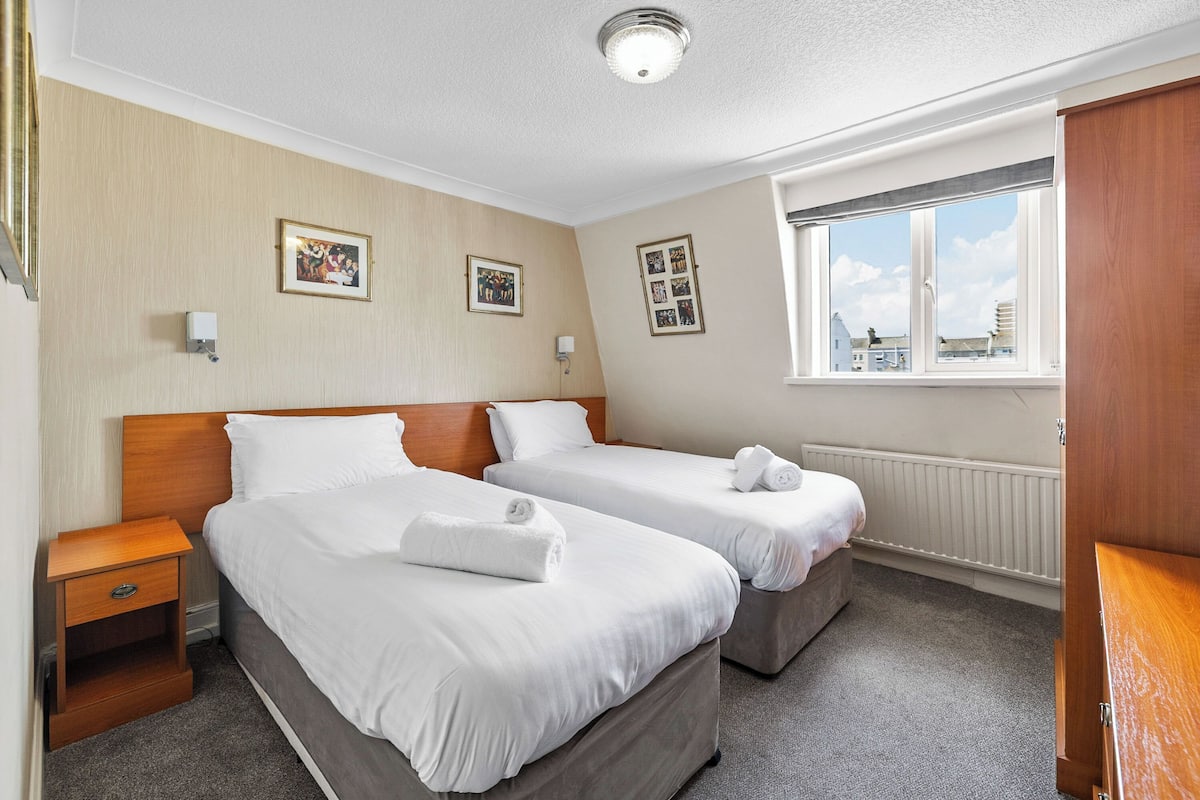 Quarto de hotel com duas camas de solteiro, mesa de cabeceira de madeira, uma mesinha, janela com vista e molduras de quadros na parede.