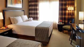 Quarto de hotel com uma cama grande, uma cama menor, uma escrivaninha com cadeira, uma luminária e um quadro na parede.