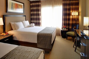 Quarto de hotel com uma cama grande, uma cama menor, uma escrivaninha com cadeira, uma luminária e um quadro na parede.