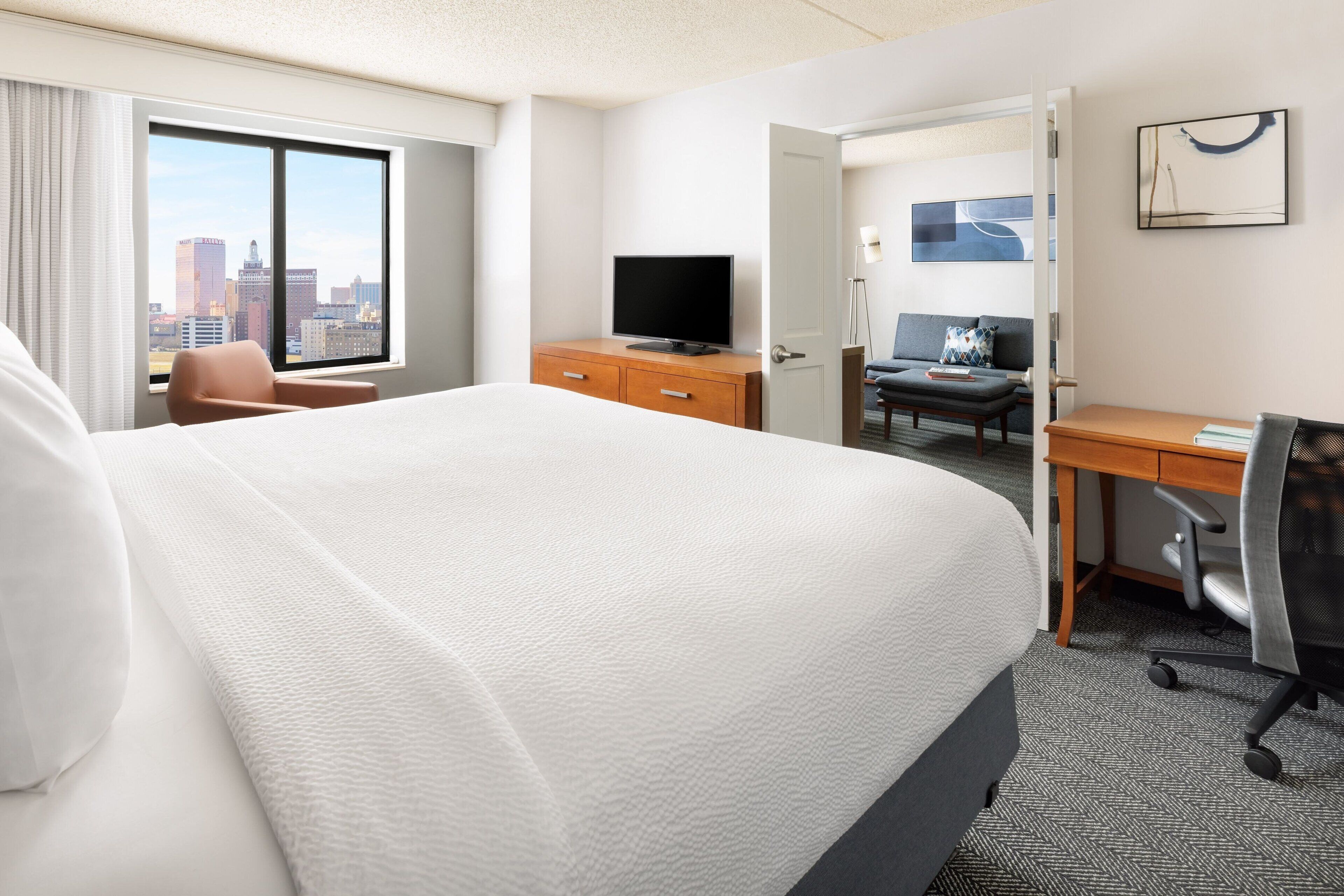 Quarto de hotel com uma cama grande, uma escrivaninha, uma cadeira, televisão e vista para a cidade através de uma janela.