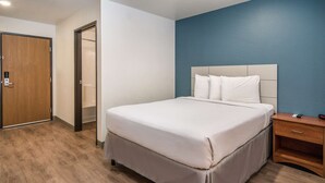 Quarto de hotel com uma cama grande, uma mesa de cabeceira de madeira e uma porta que dá para um banheiro.