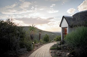Uma trilha leva a uma casa com telhado de palha, cercada por vegetação e colinas.