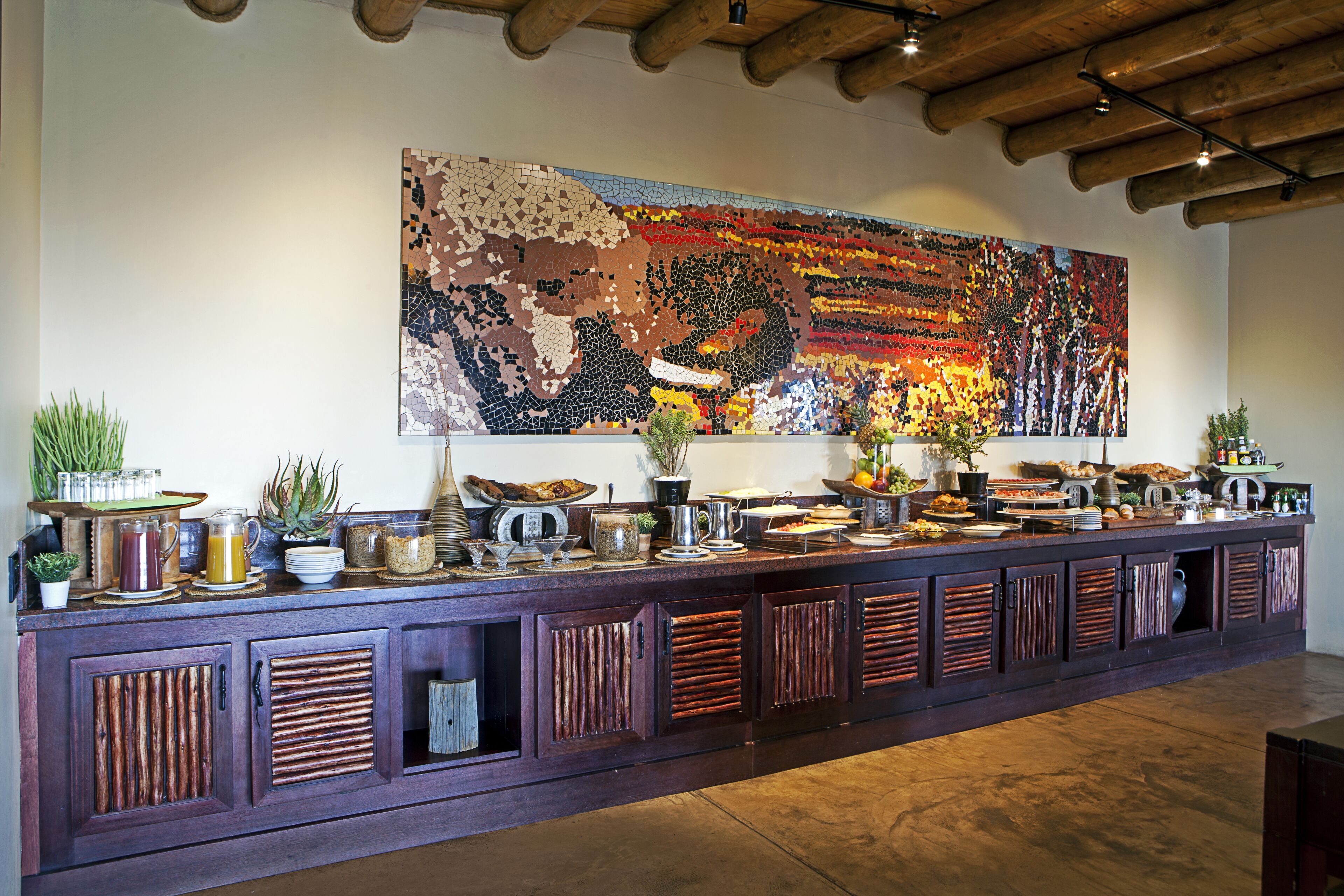 Um longo balcão de buffet com diversos alimentos e bebidas, uma grande obra de arte em mosaico na parede, e armários de madeira com design rústico.