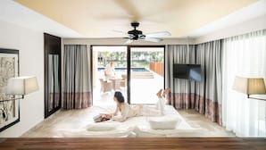 Quarto de hotel com cama, ventoinha no teto, televisão e vista para uma piscina e área de estar ao ar livre.