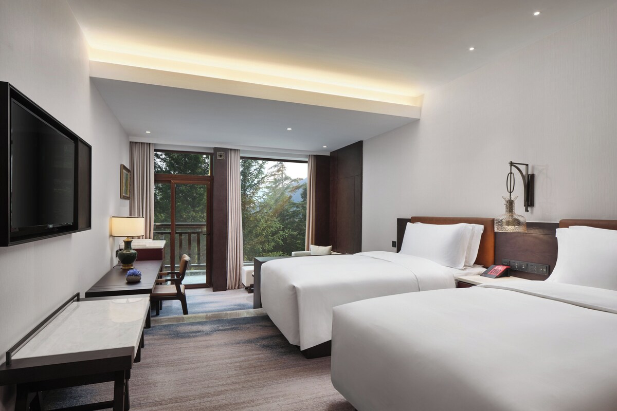 Quarto de hotel com duas camas, uma mesa, uma cadeira, uma televisão e uma janela com vista para árvores.