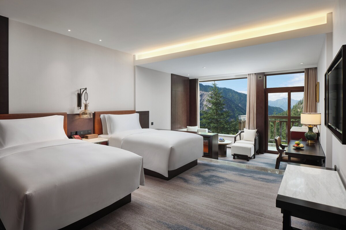 Quarto de hotel com duas camas, uma mesa, uma cadeira e vista para as montanhas.