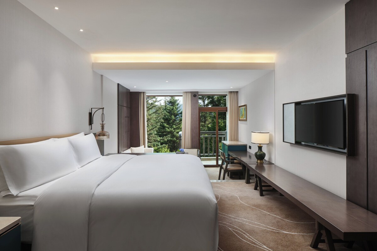 Quarto de hotel moderno com uma cama grande, uma escrivaninha, televisão de tela plana e varanda com vista para árvores.