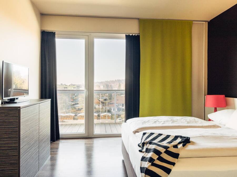 Quarto de hotel com cama, televisão em uma cômoda e vista da cidade por uma janela grande.