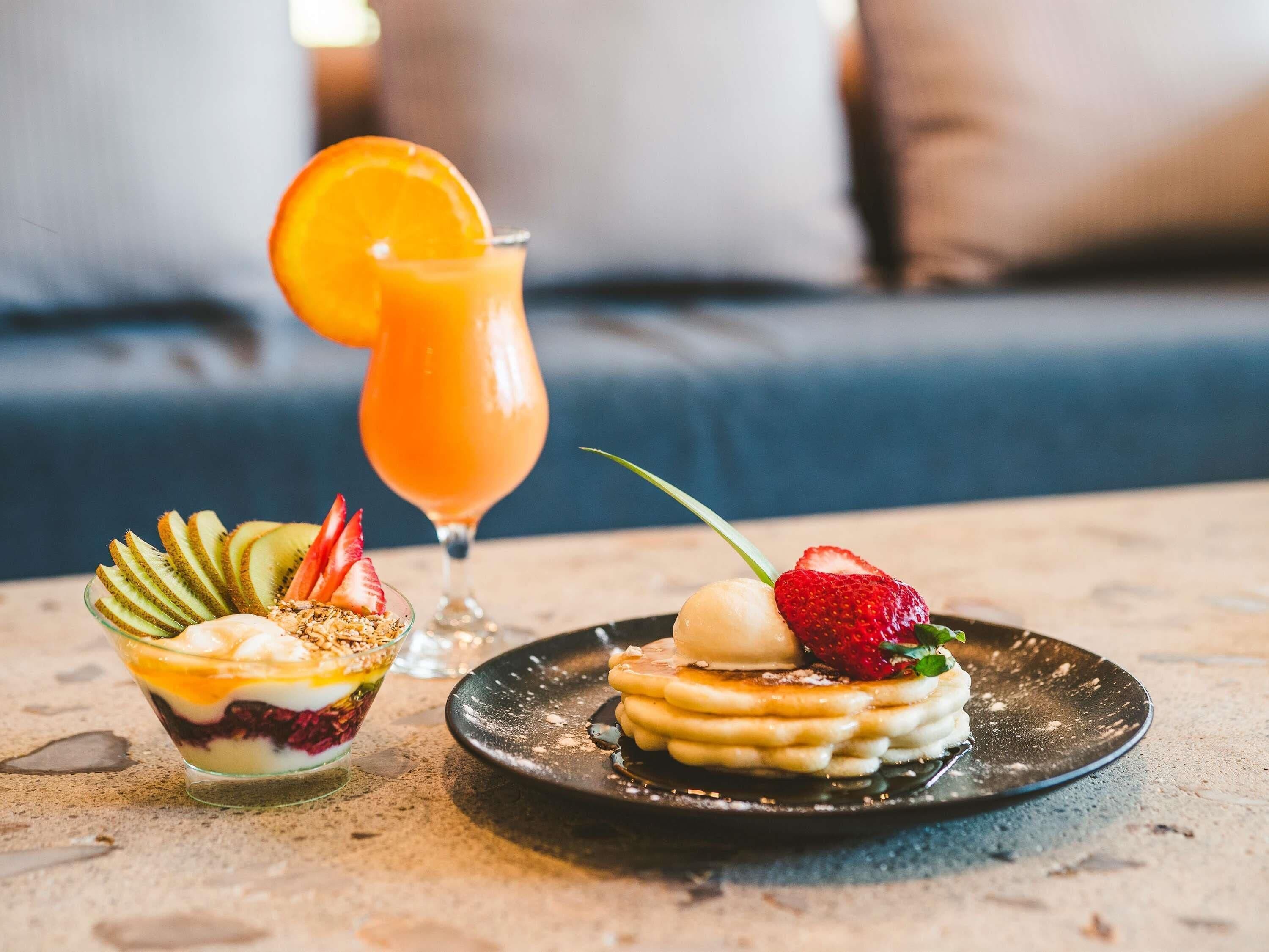 Uma bandeja de sobremesas com parfait em camadas e panquecas, um copo de suco de laranja e uma fatia de bolo com morango em um prato.