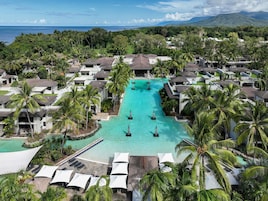 Um resort com uma piscina central, rodeado por vilas e palmeiras, com vista para o oceano e montanhas ao fundo.