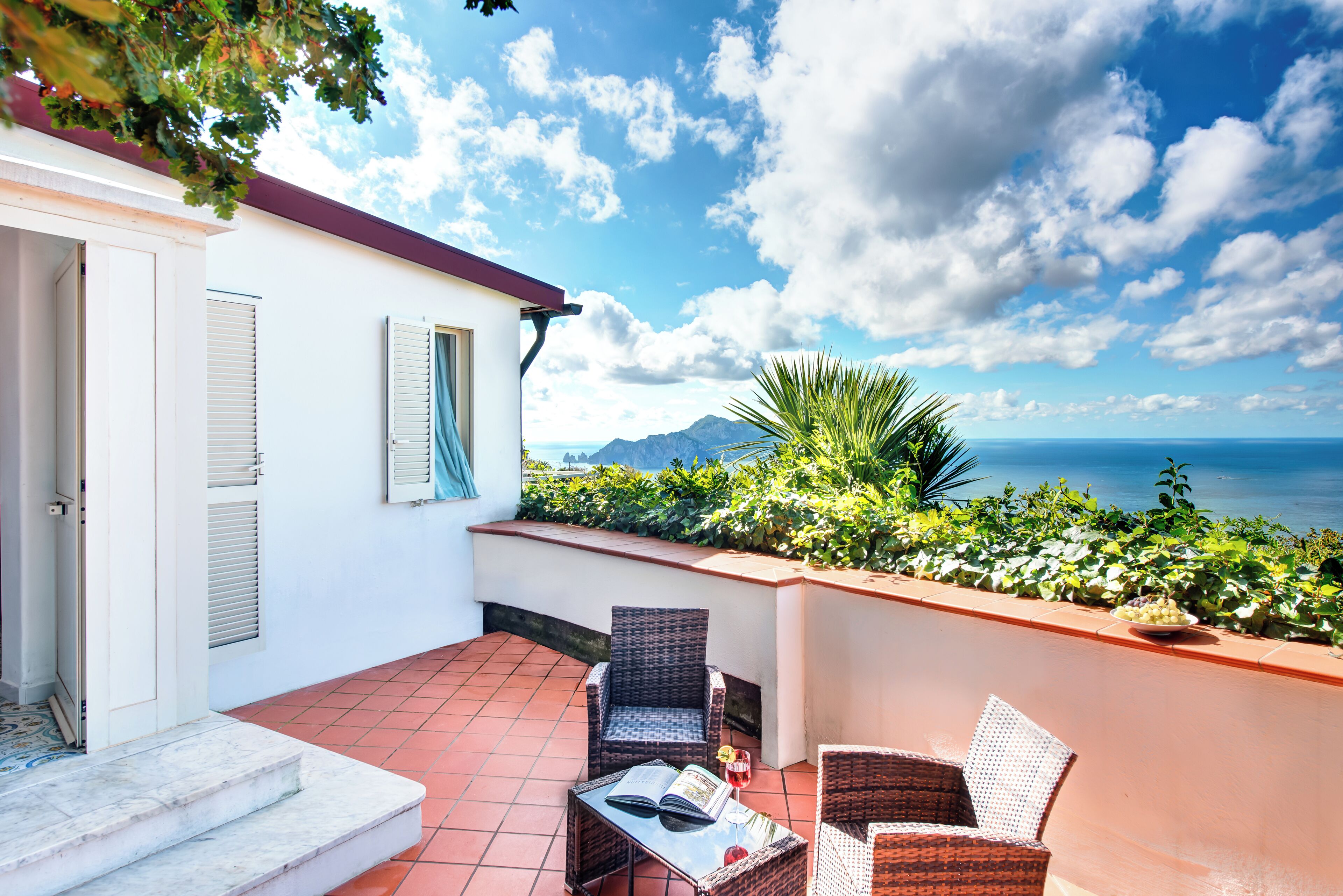 Um terraço com móveis de vime, uma mesa com um livro e um copo, e vista para o mar e montanhas.