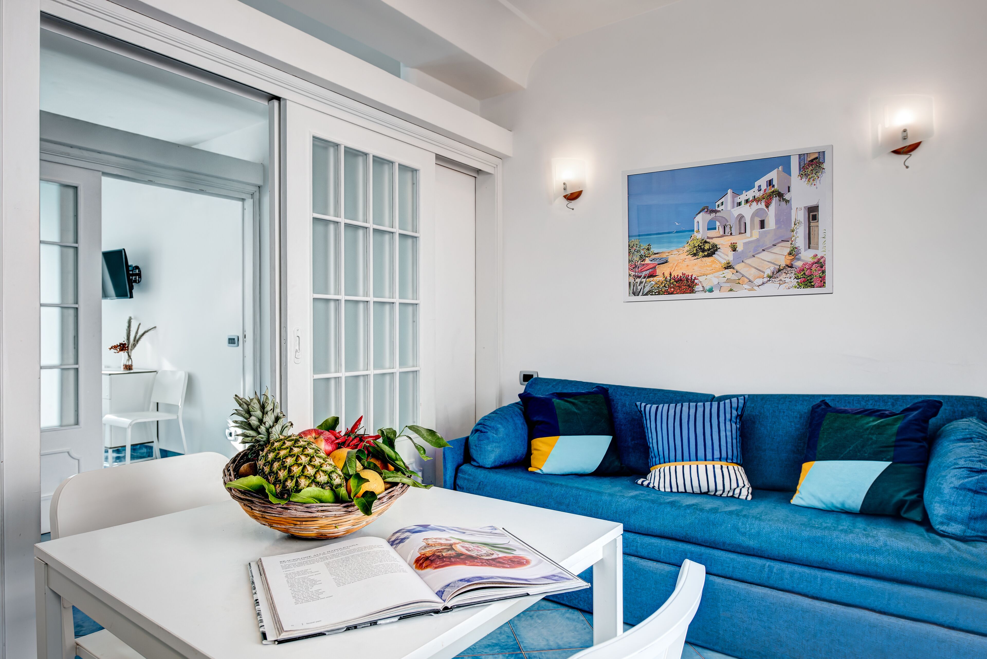 Uma sala de estar moderna com um sofá azul, uma mesa branca com uma cesta de frutas e uma revista, e um quadro com uma paisagem costeira na parede.