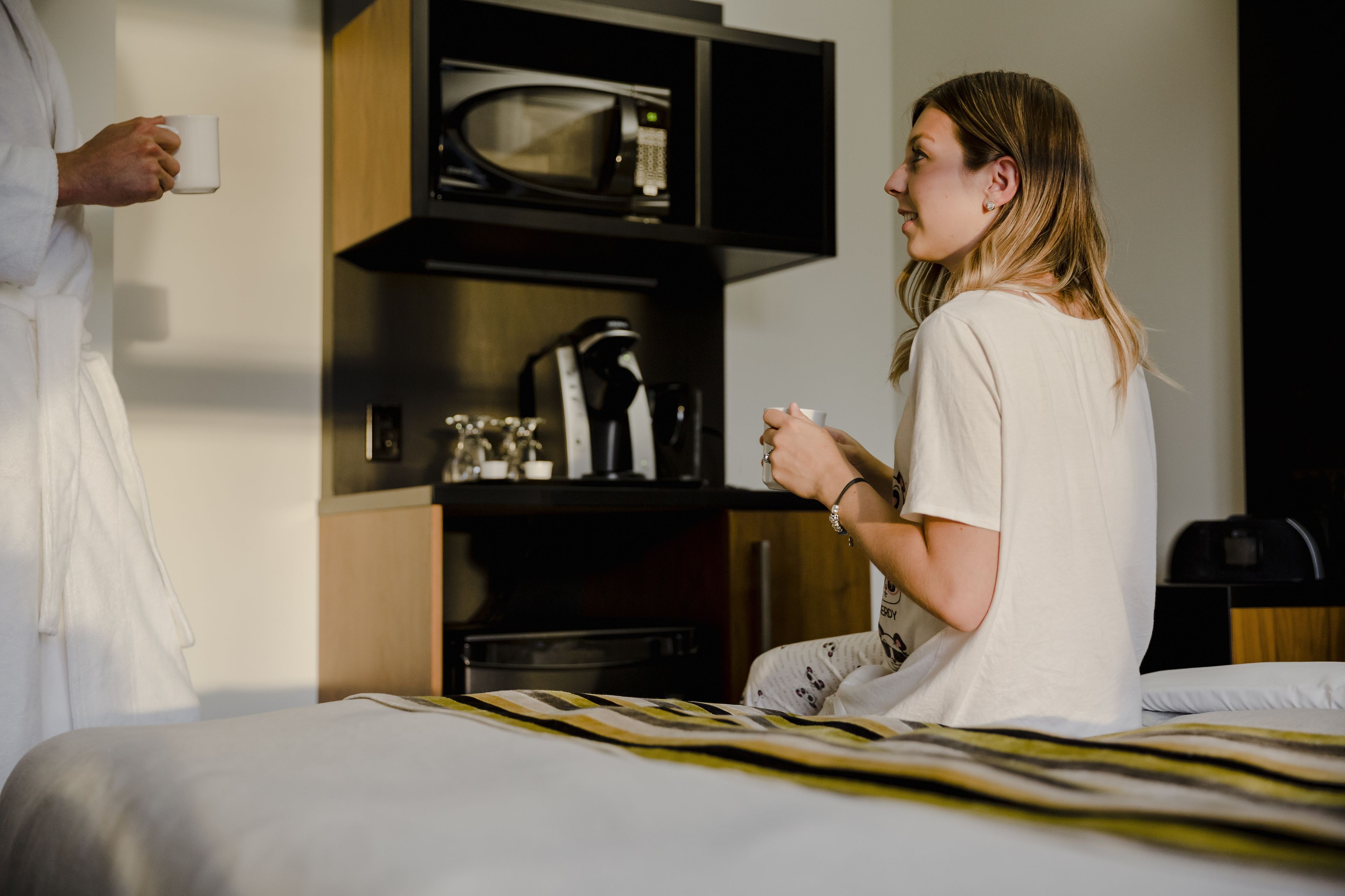 Quarto de hotel com uma cama, micro-ondas, cafeteira e uma pessoa segurando uma xícara.