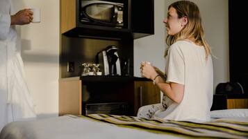 Quarto de hotel com uma cama, micro-ondas, cafeteira e uma pessoa segurando uma xícara.