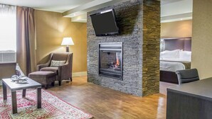 Quarto de hotel com lareira de pedra, televisão de tela plana montada acima, cama com lençóis brancos, área de estar com uma cadeira e mesa, e um tapete vermelho estampado.
