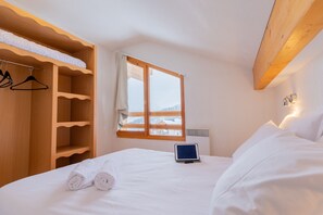 Cama bem arrumada com um laptop, guarda-roupa de madeira e janela com vista para uma paisagem nevada.