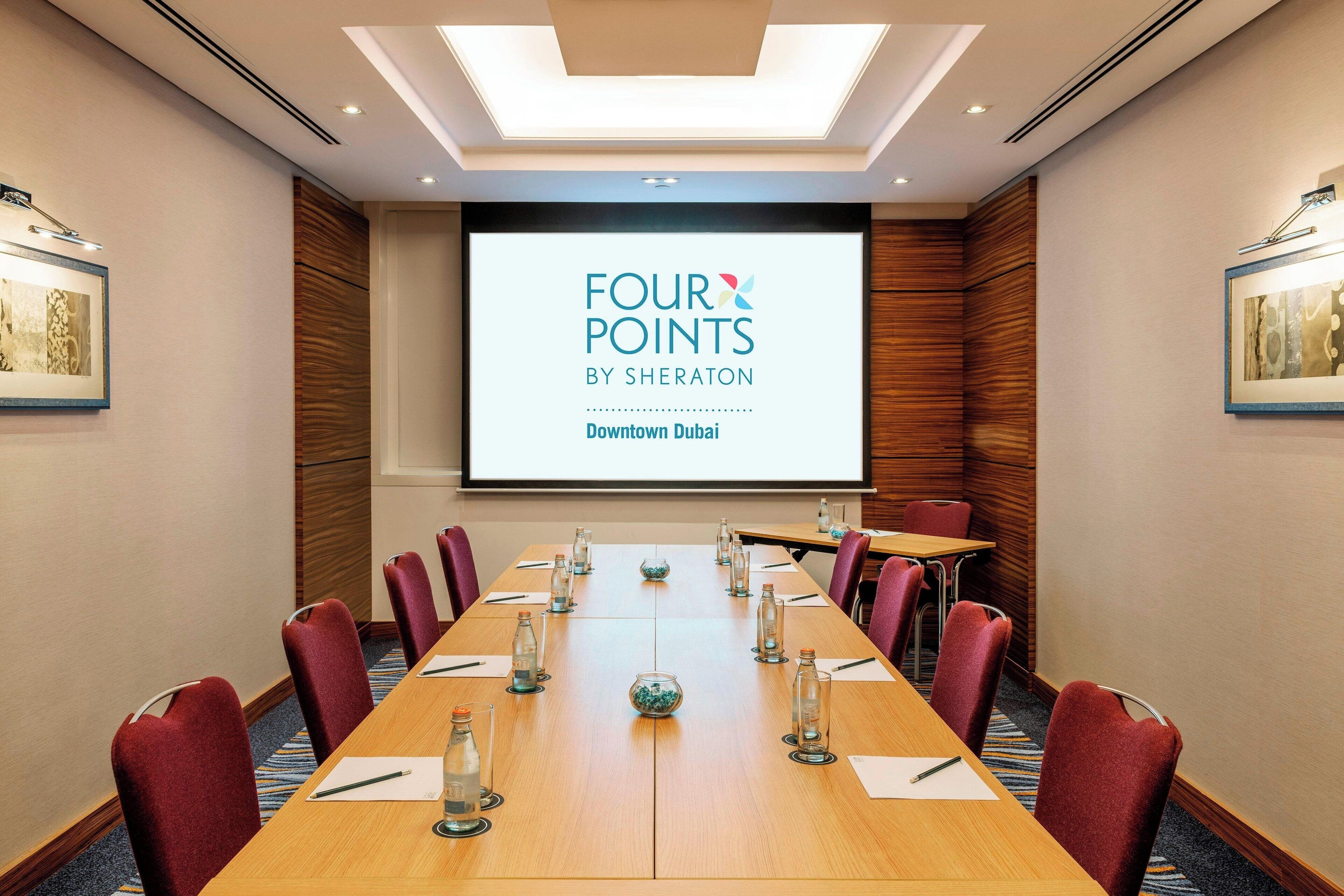 Uma sala de conferências com uma mesa comprida, cadeiras vermelhas e uma tela exibindo "FOUR POINTS BY SHERATON Downtown Dubai".