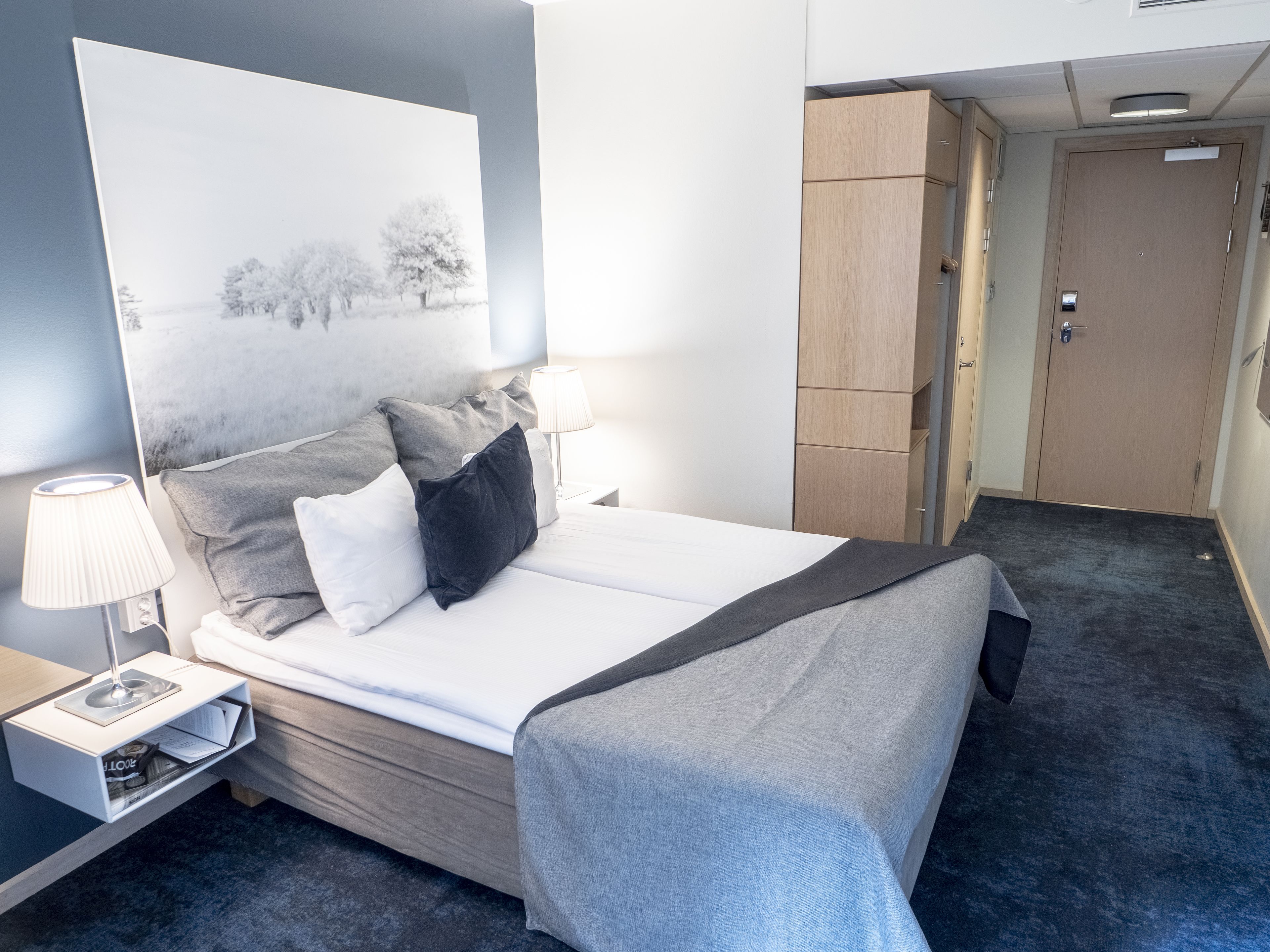 Quarto de hotel moderno com cama, mesa de cabeceira, abajur e uma grande pintura na parede.