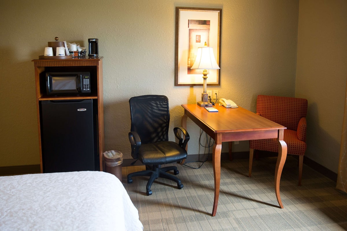 Quarto de hotel com cama, escrivaninha com luminária, cadeira, micro-ondas e lixeira.