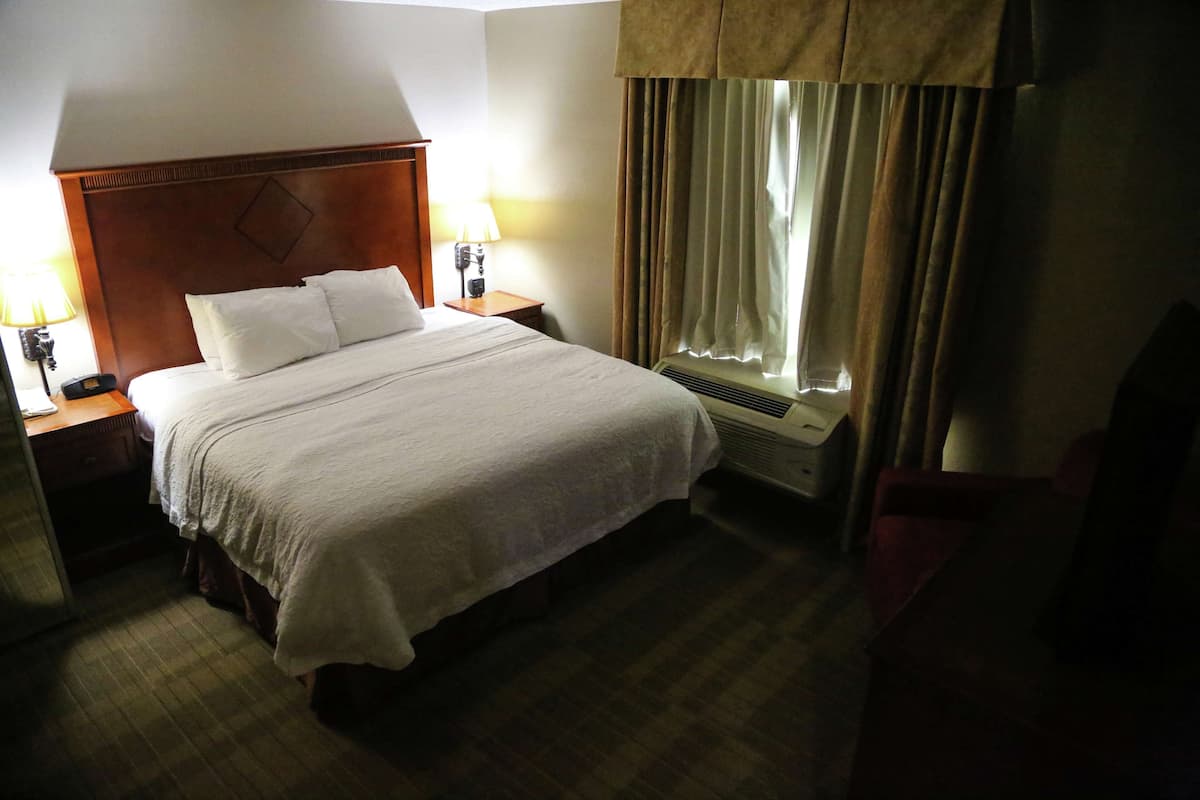 Quarto de hotel com uma cama grande, uma mesa de cabeceira com abajur, aquecedor e uma janela com cortinas.