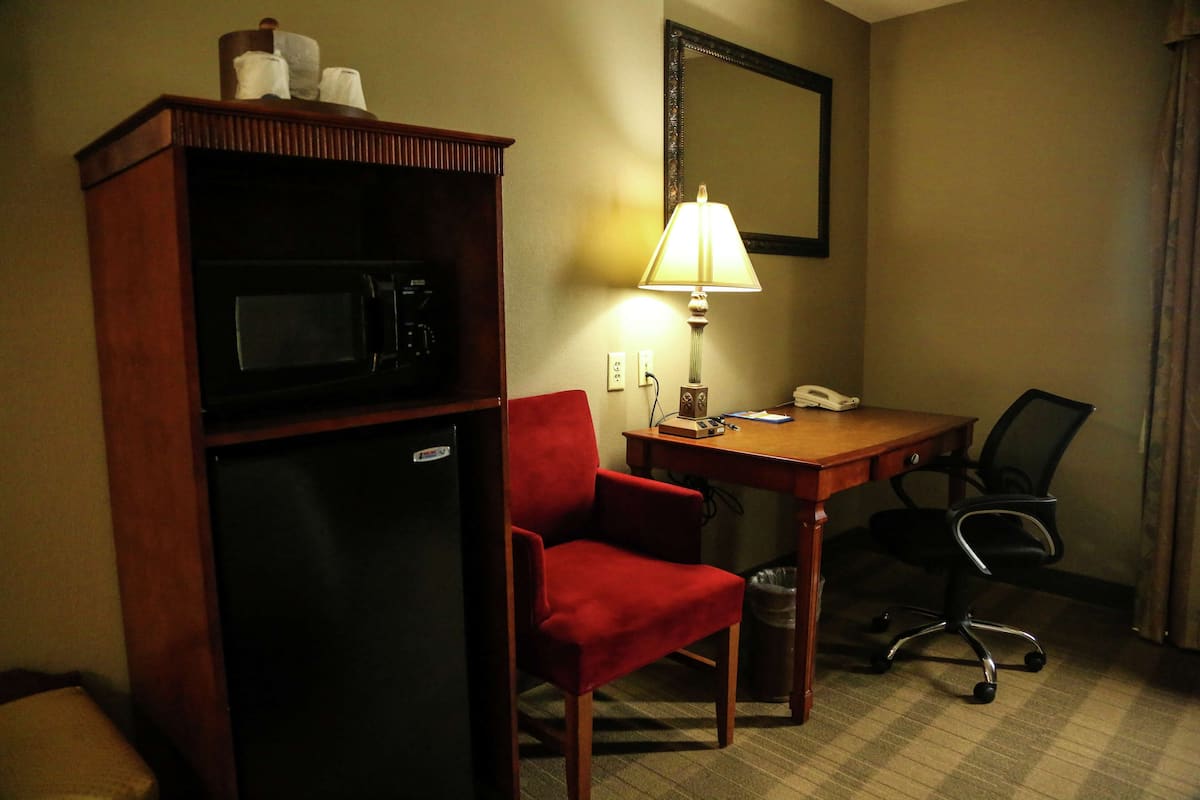 Quarto de hotel com cozinha compacta, escrivaninha com luminária, uma cadeira vermelha e uma cadeira de escritório preta.
