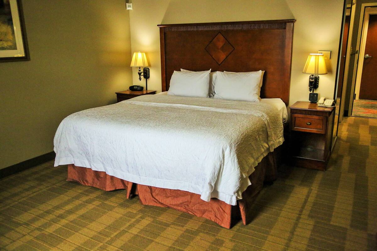 Quarto de hotel com uma cama grande, duas mesas de cabeceira com abajures, um painel de cabeceira em madeira e piso xadrez.