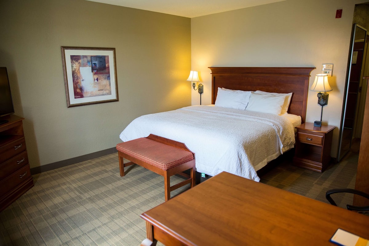 Quarto de hotel com cama, mesas de cabeceira, escrivaninha, cadeira, banqueta e um quadro na parede.