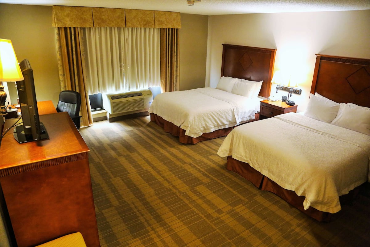 Quarto de hotel com duas camas, uma mesa de trabalho com luminária, uma cadeira e uma janela com cortinas.