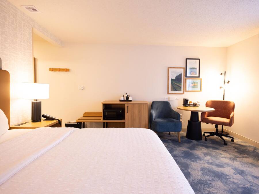 Quarto de hotel com cama, escrivaninha, duas cadeiras, micro-ondas e uma pequena mesa com abajur.