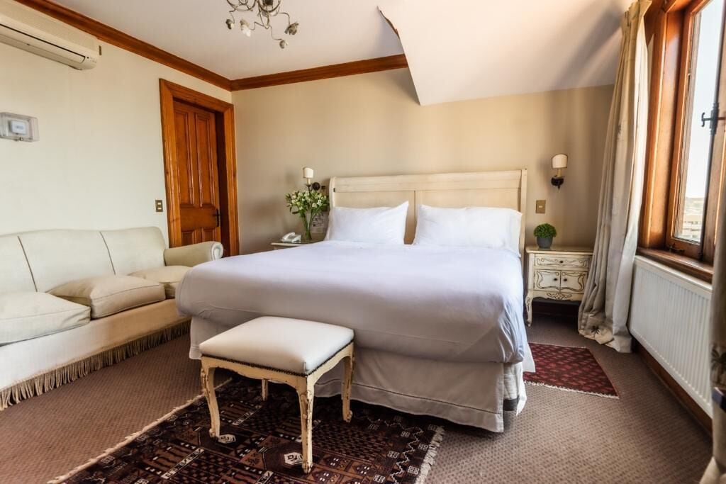 Quarto com uma cama grande, uma mesa de cabeceira, uma luminária e uma cadeira, além de uma janela com cortinas.