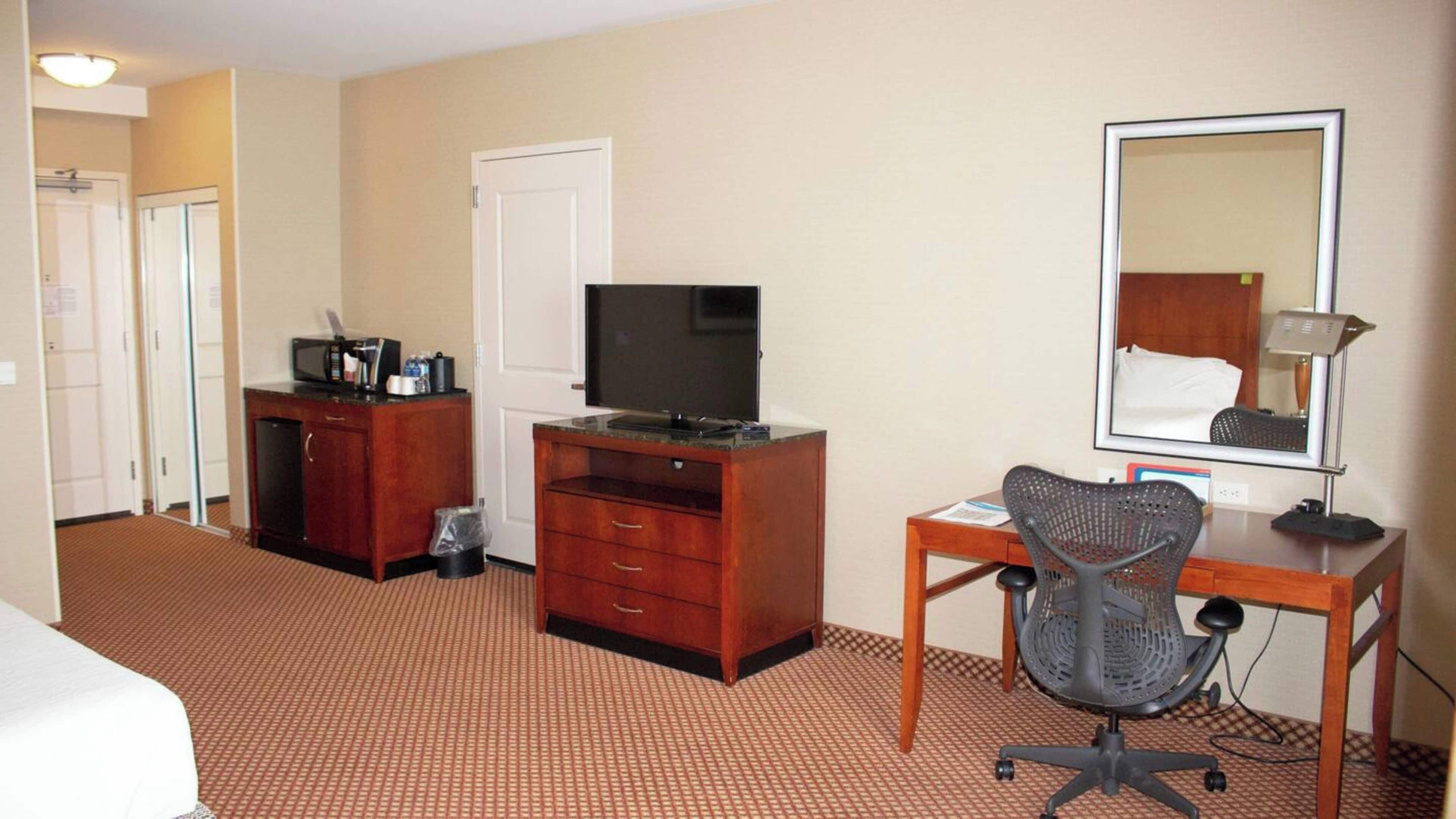 Quarto de hotel com uma cama, uma escrivaninha de madeira com um computador, uma cadeira, uma televisão, um espelho e um guarda-roupa.